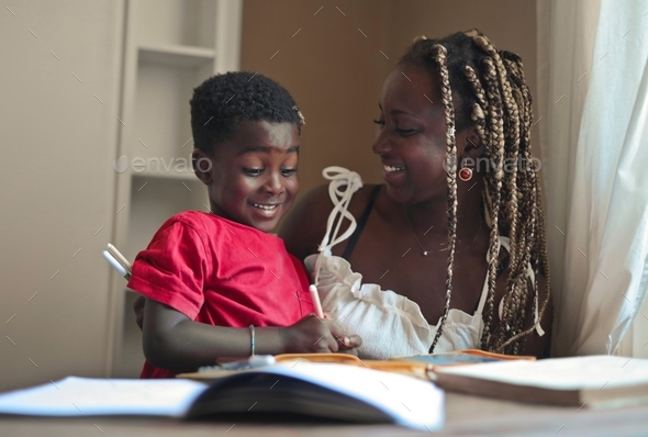 Portrait of a black kid doing his homework with his mother in front of ...