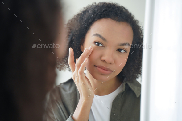 Mixed race girl touches delicate under eye skin, applying facial cream ...