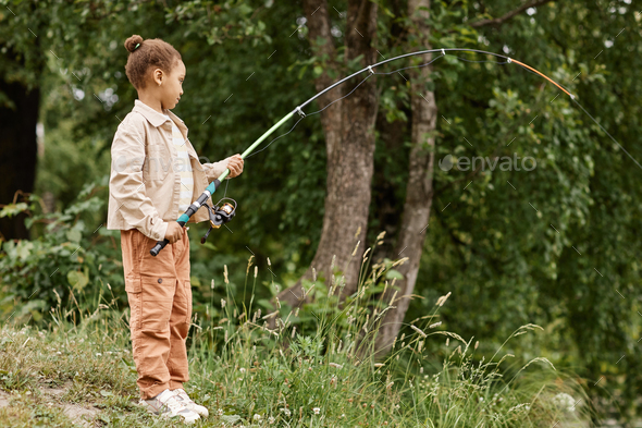 Side view black little girl fishing by river with fishing rod bending ...