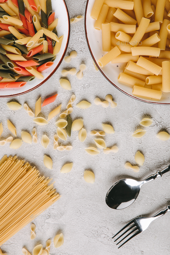 top view of various raw pasta and cutlery on messy concrete tabletop ...