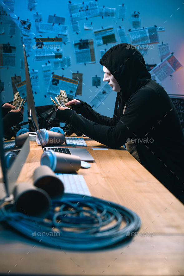 side view of hacker in mask counting stolen money at his workplace ...