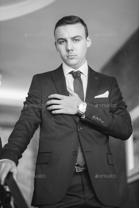 Grayscale of a confident Caucasian young man in a blue suit leaning on ...