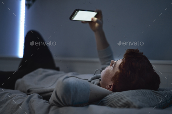 Cheerful caucasian teenage boy watching mobile phone while lying in bed ...