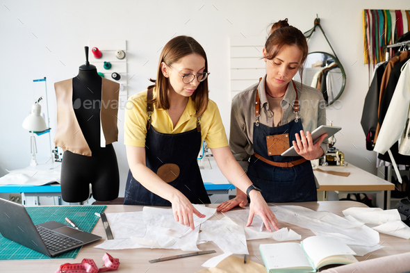 Two female tailors choosing sewing paper patterns by workplace in ...