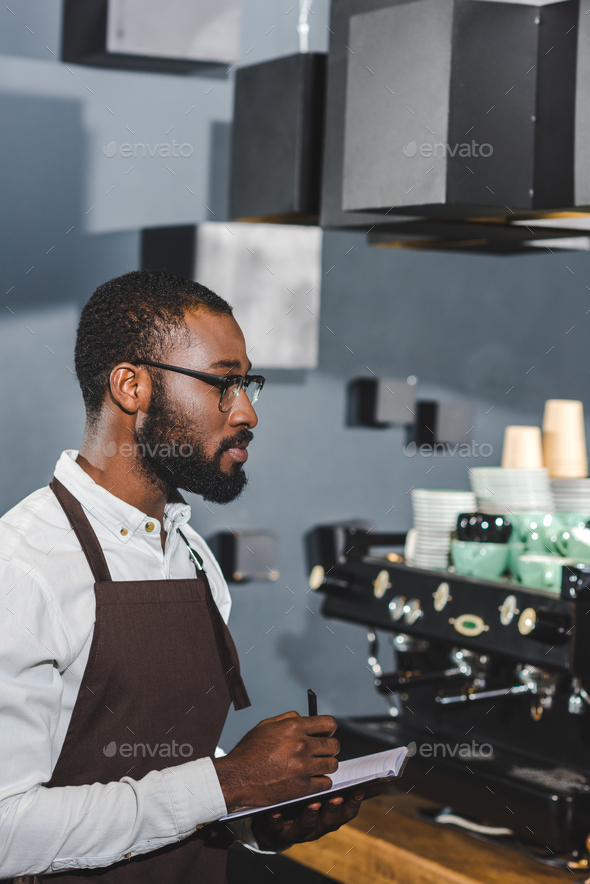 side view of young african american barista in eyeglasses taking notes ...