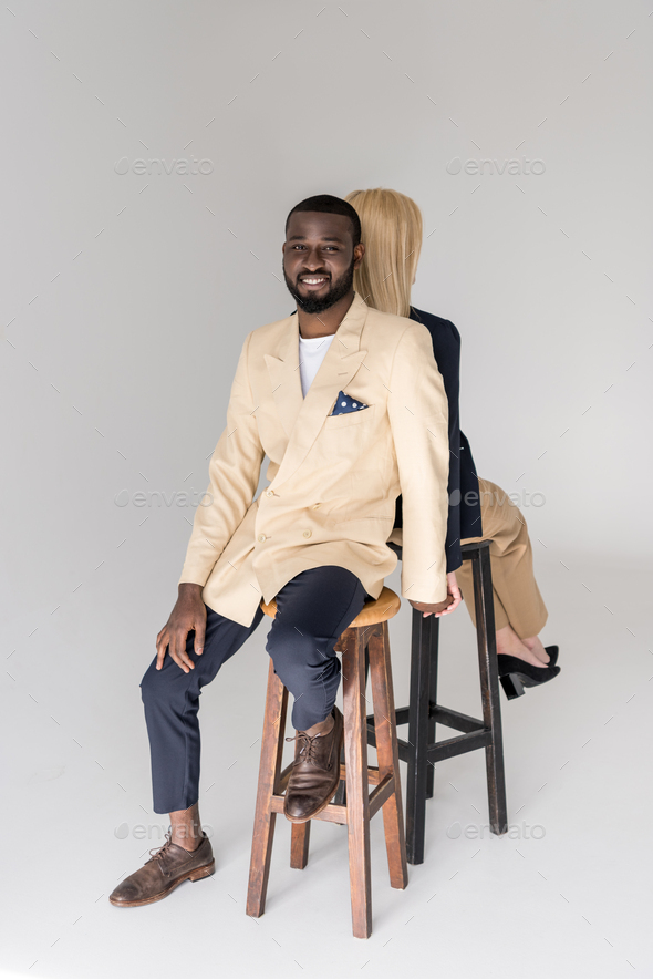 stylish young multiethnic couple sitting back to back, man smiling at ...