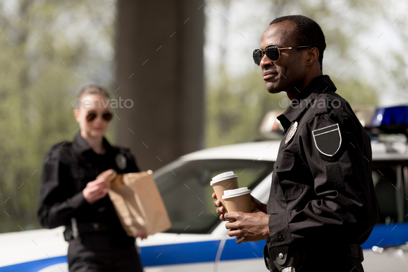 young police officers with coffee to go and paper bag with lunch having ...