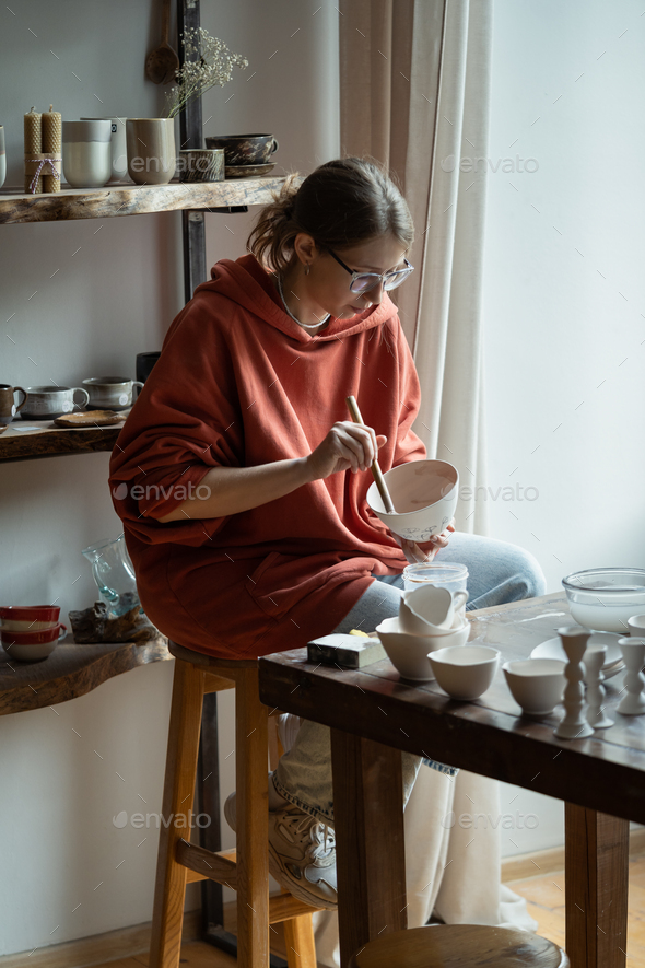 Focused young woman sits in pottery workshop working on making crockery ...