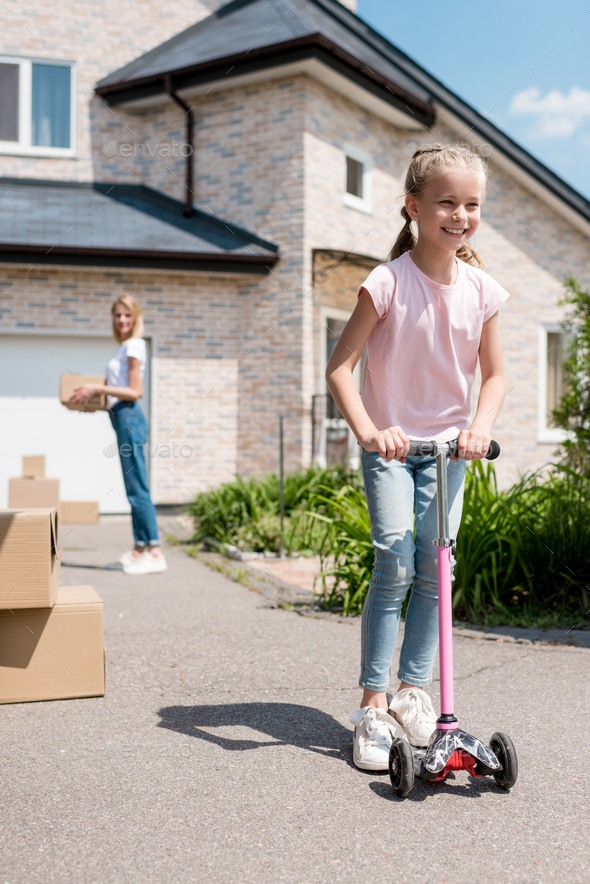 smiling little child riding on kick scooter while her mother unpacking ...