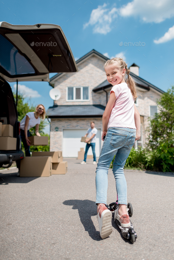 happy little kid riding on kick scooter and her parents unpacking ...
