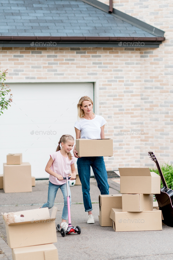 mother unpacking cardboard boxes and little daughter riding on kick ...