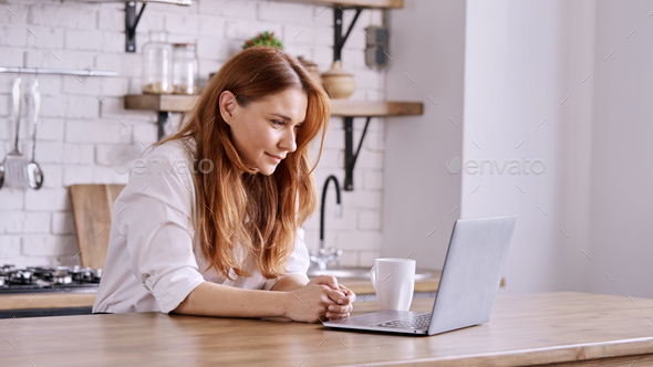 Caucasian female working on her laptop and drinking coffee in a kitchen ...