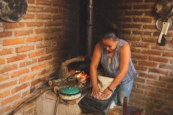 Mexican female preparing corn mace on a metate and a wood stove to make ...