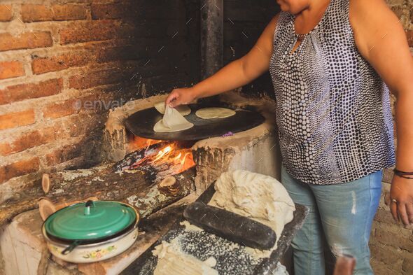 Mexican female preparing corn mace on a metate and a wood stove to make ...