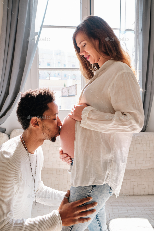 Man kissing his pregnant wife belly at home. Stock Photo by alvarogonzalez