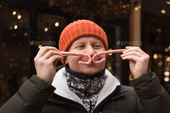 man in hat and winter clothes makes mustache from candy canes, posing for photo at Christmas ...