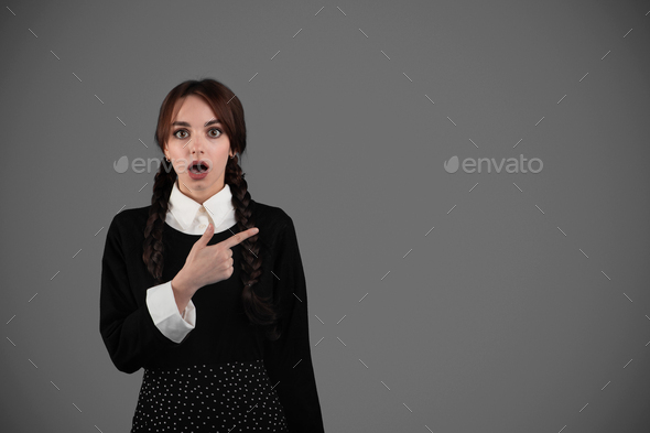 Shocked young female student in gothic black clothes with pigtails with open mouth point finger ...