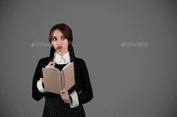 Pensive smart young girl student in gothic black clothes with pigtails with book, got idea and ...