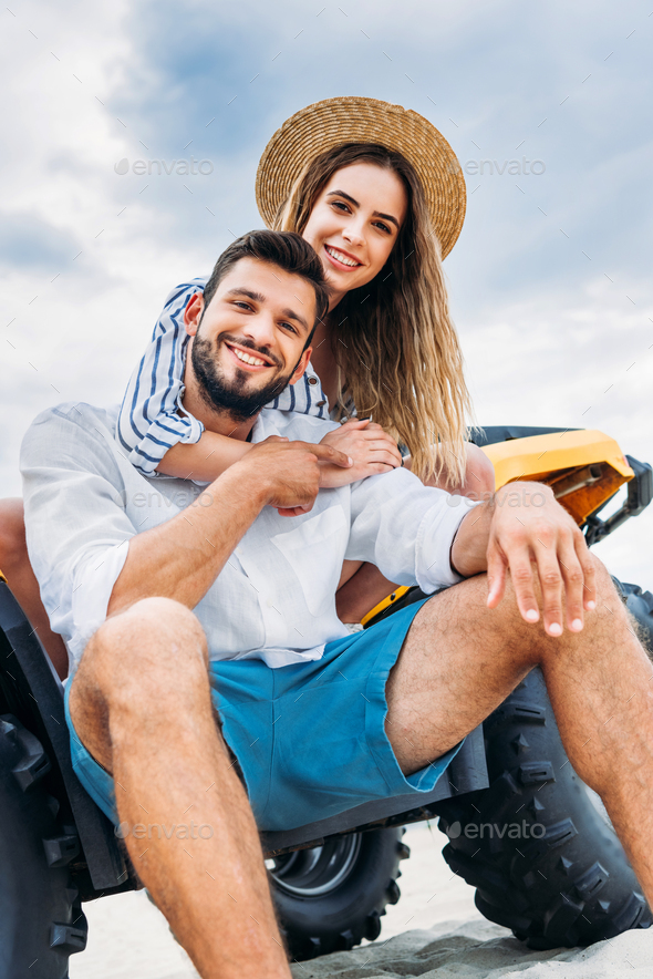 bottom view of happy young couple sitting on ATV in front of cloudy sky ...