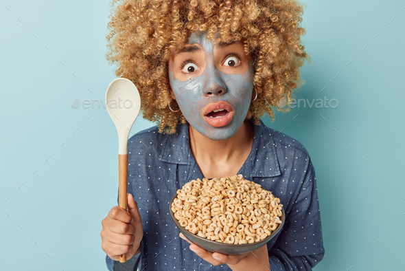Scared frightened curly haired woman holds spoon and bowl of cereals ...