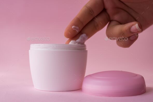 Closeup shot of a hand dipping into a container of skin cream on a pink ...