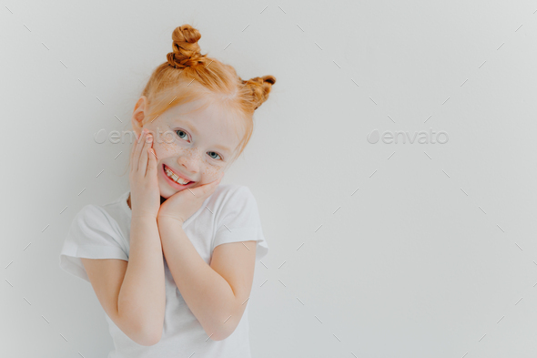 Happy small redhead girl tilts head, keeps hands on cheeks, smiles ...