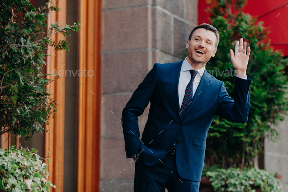 Shot of cheerful male banker in luxury black suit, waves with hand as ...
