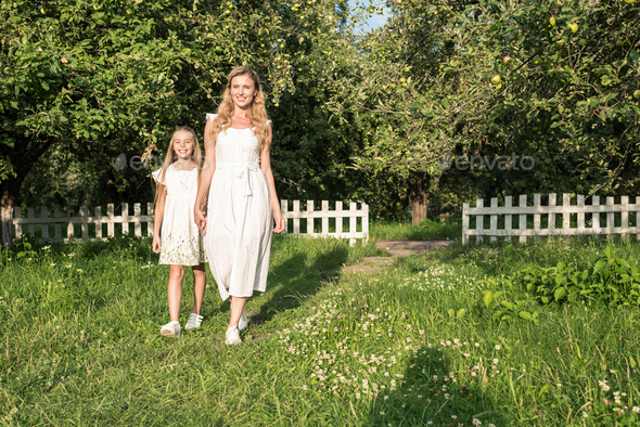 beautiful mother and daughter in dresses holding hands in orchard with ...