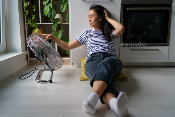 Pleased Asian woman blown by electric fan sits on floor enjoy cool wind ...