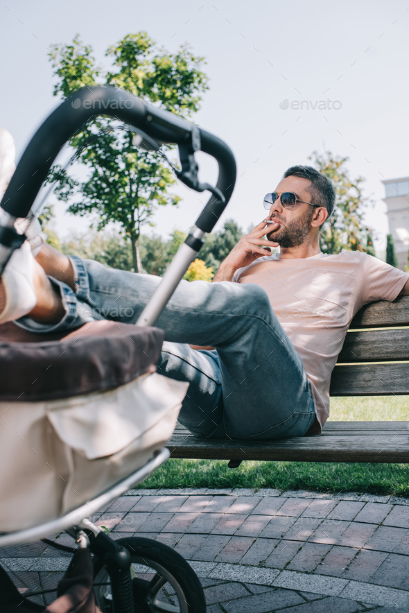 father smoking cigarette on bench with legs on baby carriage in park ...