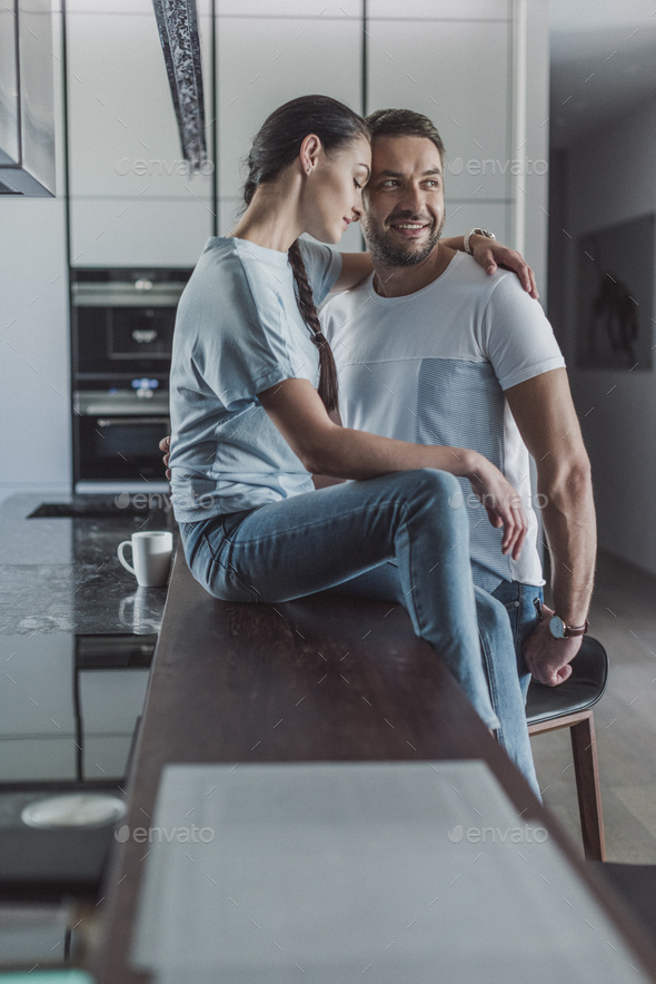 side view of attractive woman sitting on kitchen counter and embracing ...