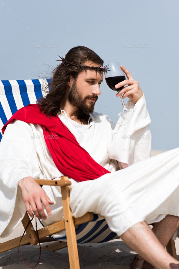pensive Jesus in robe and red sash sitting on sun lounger with glass of ...