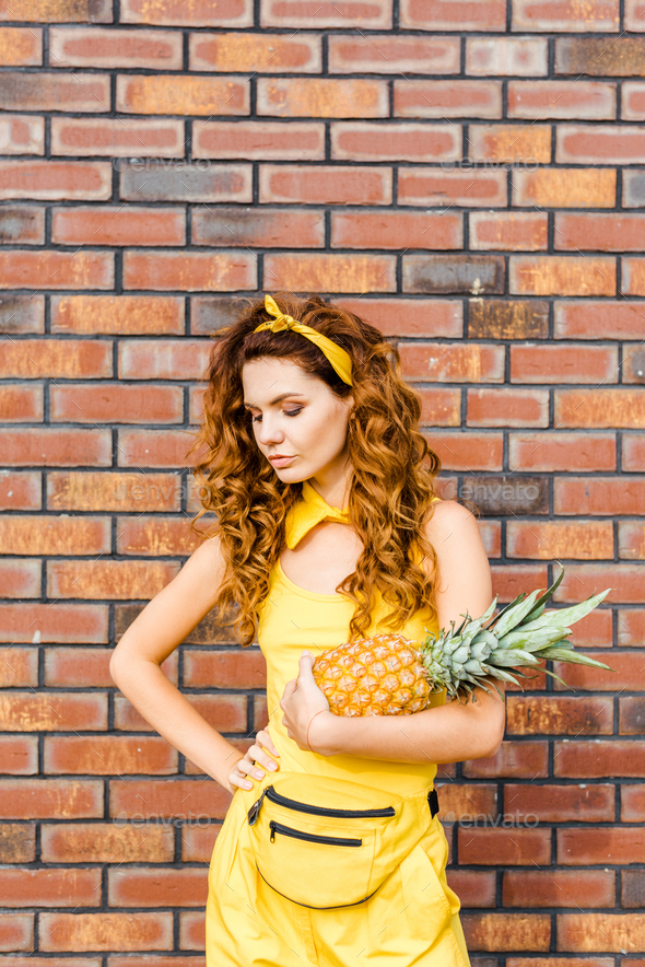 beautiful young woman in yellow clothes holding pineapple in front of brick wall Stock Photo by ...