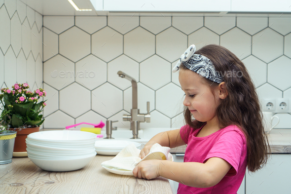 Happy little girl wiping clear plates with dry towel on kitchen set ...