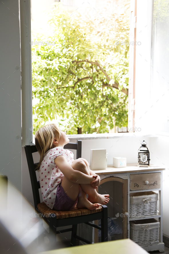 Little girl sitting on chair looking out of window Stock Photo by westend61
