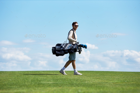 Minimal side view rich man carrying golf bag walking on green field ...
