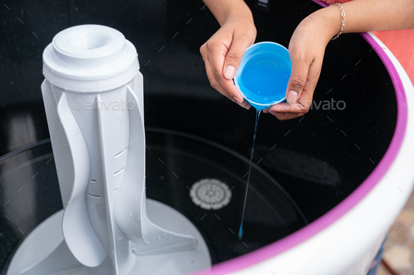 A young woman is pouring some liquid detergent into a top load washing ...