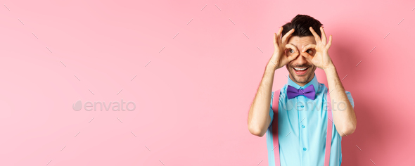 Cheerful man in bow-tie and suspenders looking through hand binoculars ...
