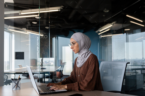 Serious and thinking woman in hijab working inside office with laptop ...