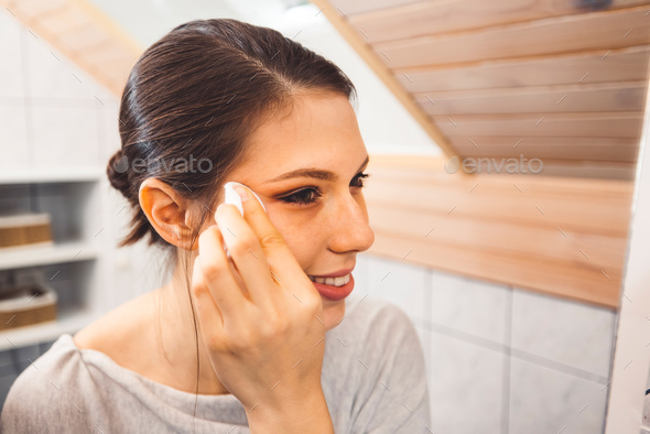 Smiling cheerful young woman cleaning make up off her face late at ...