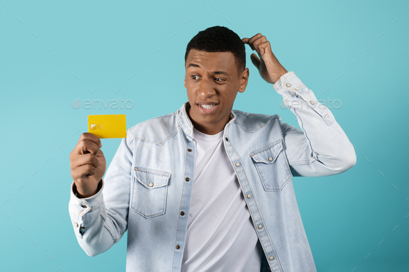 Pensive millennial black guy scratching head, look at credit card ...