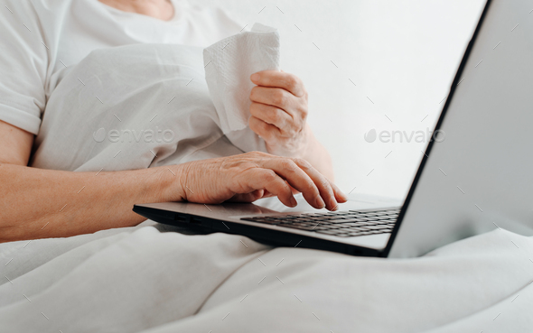 Elderly sick woman working on laptop while sitting on bed in bedroom ...
