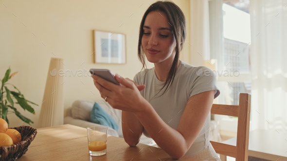 Smiling young woman scrolling through photo on her mobile phone while sitting in the kitchen ...