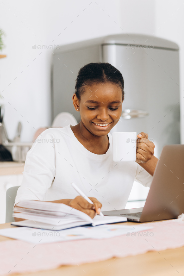 Young black woman sitting in the kitchen writes something and working ...