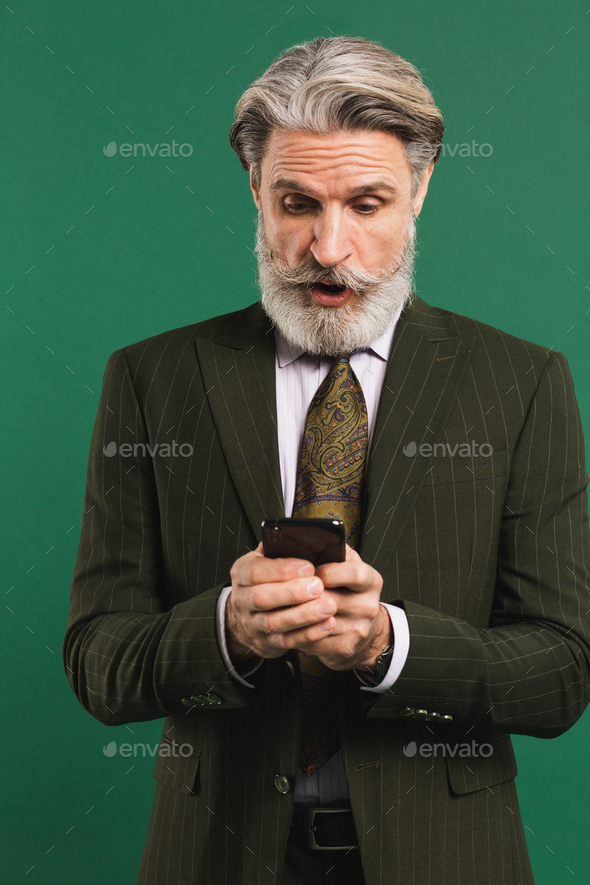 Bearded middle-aged man in khaki suit looks at phone and shows shock on ...