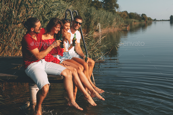 Happy group of young people drinking beer on a dock by the river during ...