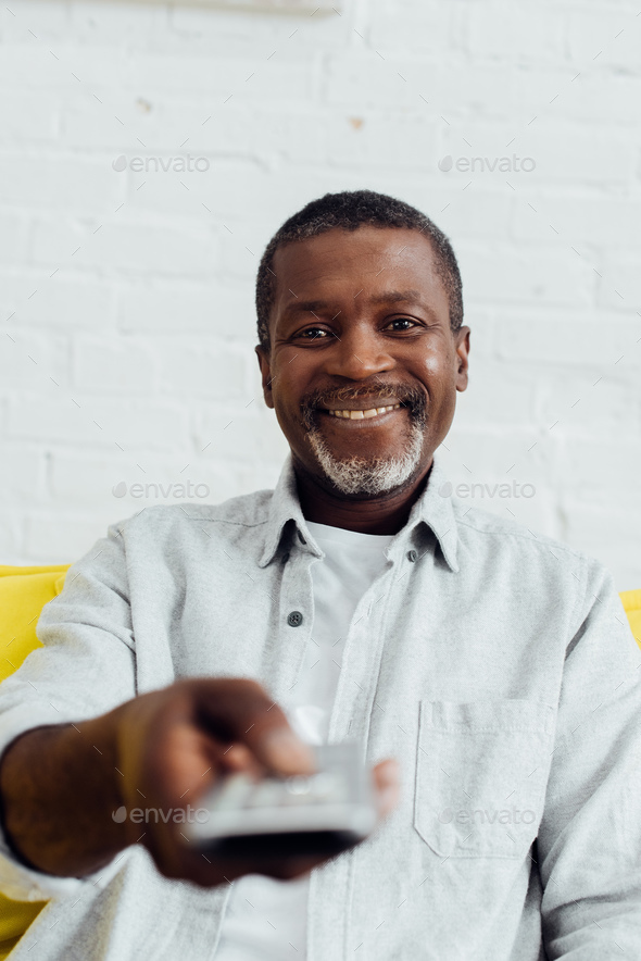 smiling handsome african american man sitting on sofa with tv remote ...