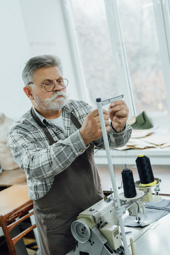 focused middle aged tailor in apron cutting strings on sewing machine ...