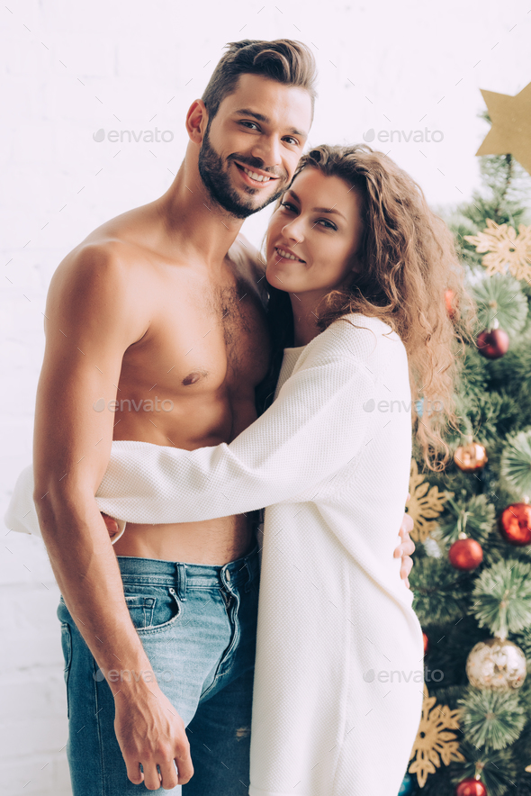 curly smiling woman hugging shirtless muscular boyfriend near christmas ...