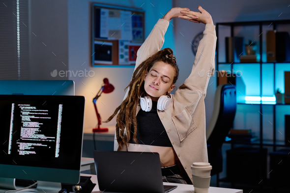 Young tired female programmer with headphones on neck stretching arms ...